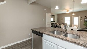Uma cozinha moderna com bancadas de granito, pia de aço inoxidável e máquina de lavar louça. Há uma área de jantar com mesa e cadeiras ao fundo.