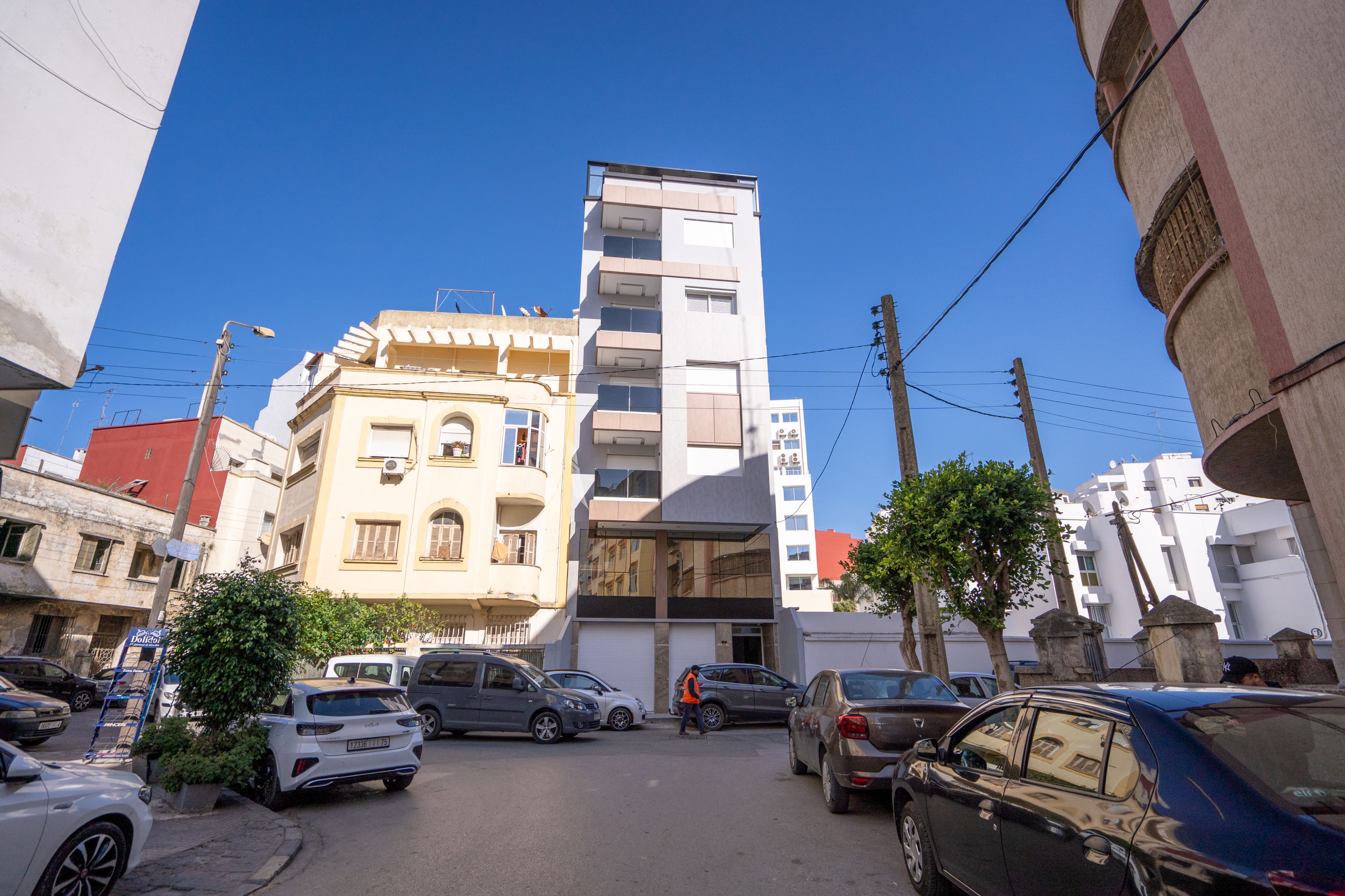 Vista da rua com carros estacionados, um prédio com vários andares e um céu azul límpido.