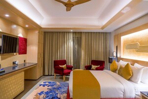 Quarto de hotel com cama, duas poltronas vermelhas, televisão, escrivaninha e uma janela grande com cortinas.