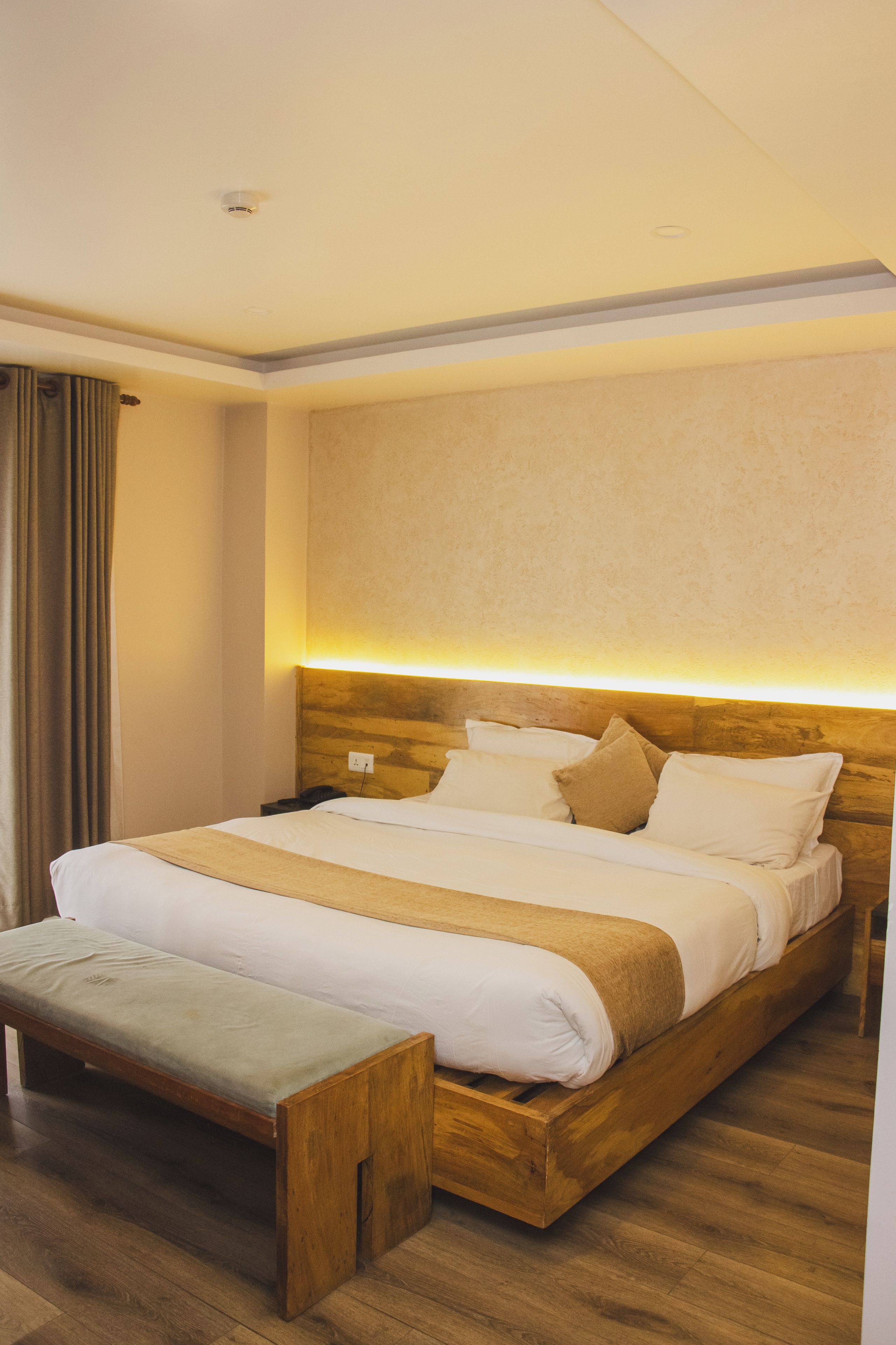 Quarto de hotel com cama de madeira, um banquinho aos pés da cama e cabeceira com iluminação integrada.