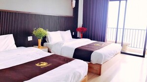 Quarto de hotel com duas camas, uma cabeceira de madeira escura, uma janela com cortinas e uma pequena mesa com um vaso de flores.