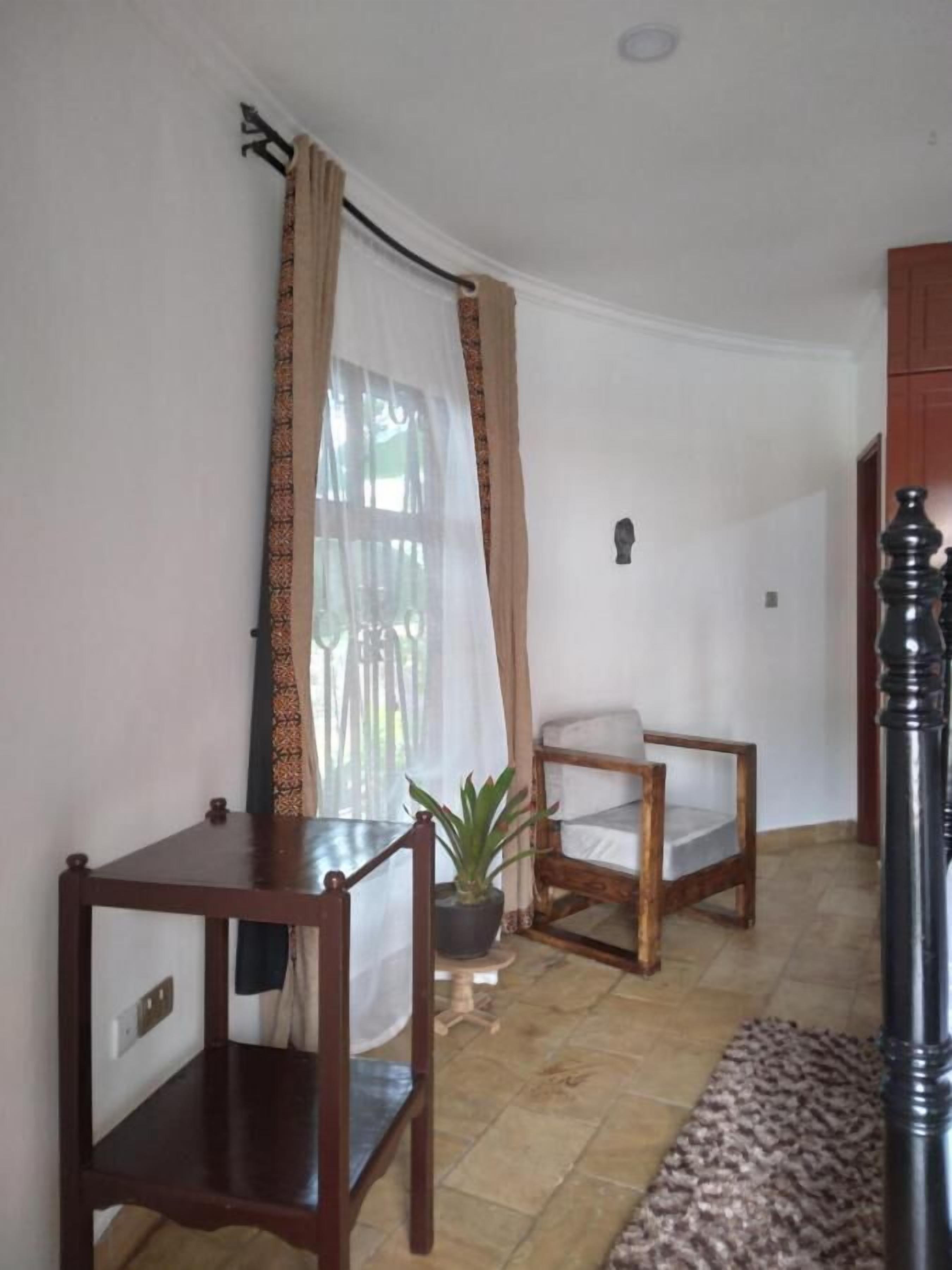 Quarto com mesa de madeira e duas cadeiras de madeira, uma planta em vaso e vista do exterior através de cortinas translúcidas.