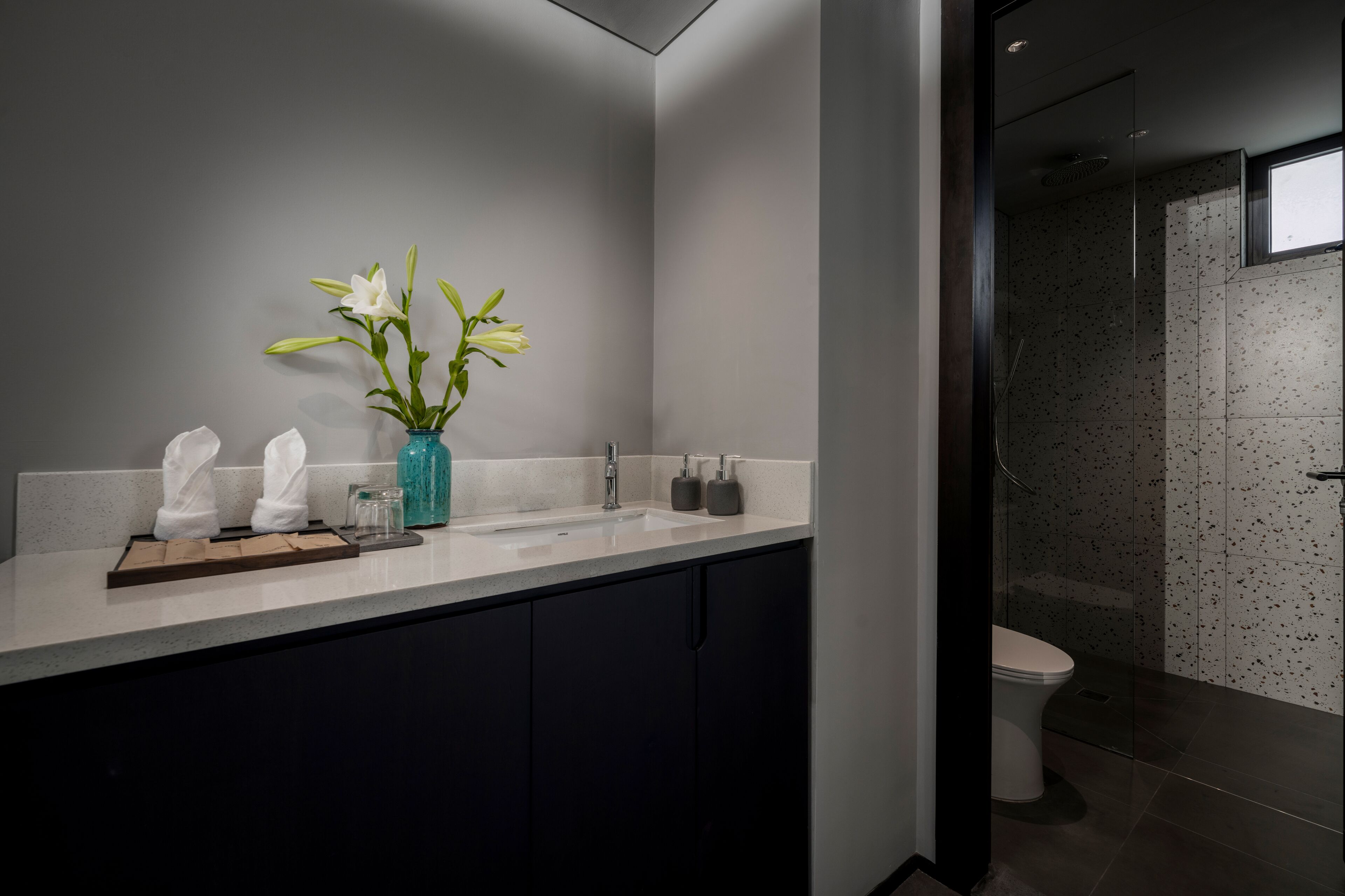 Uma casa de banho moderna com bancada branca, um vaso azul com flores brancas, uma pia e uma sanita.
