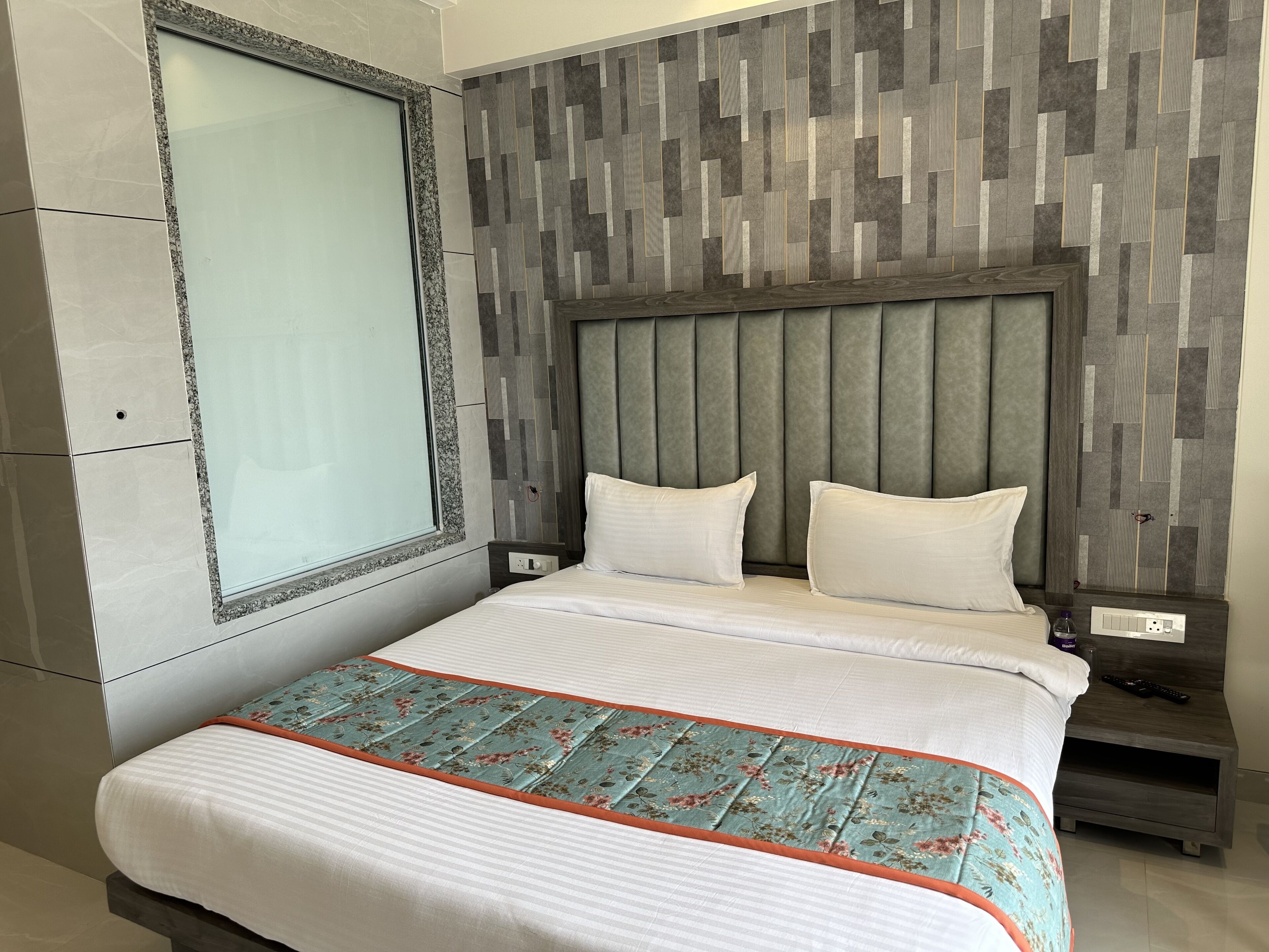 Quarto de hotel moderno com uma cama grande, cabeceira de madeira, mesa de cabeceira e parede com padrão geométrico.