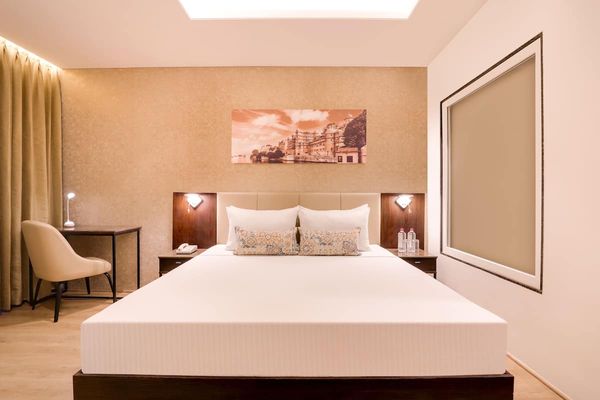 Quarto de hotel com uma cama grande, uma escrivaninha, uma cadeira, mesa de cabeceira com abajur e um quadro na parede.