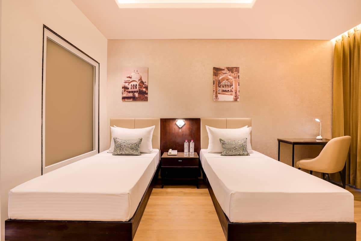 Quarto de hotel com duas camas de solteiro, uma mesa de cabeceira com telefone e garrafas de água, uma escrivaninha com cadeira e duas luminárias fixadas na parede.