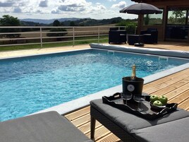 Uma área à beira da piscina com um deck de madeira, uma espreguiçadeira, uma mesa com champanhe, copos e uma tigela de maçãs.