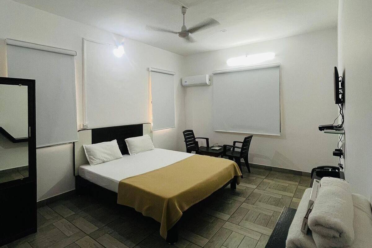 Quarto de hotel com cama, escrivaninha, cadeira, espelho, ventoinha no teto e ar condicionado.