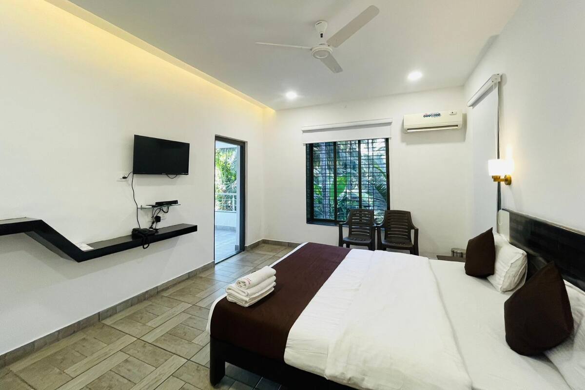 Quarto de hotel com cama, televisão, janela, ventoinha no teto e ar condicionado.