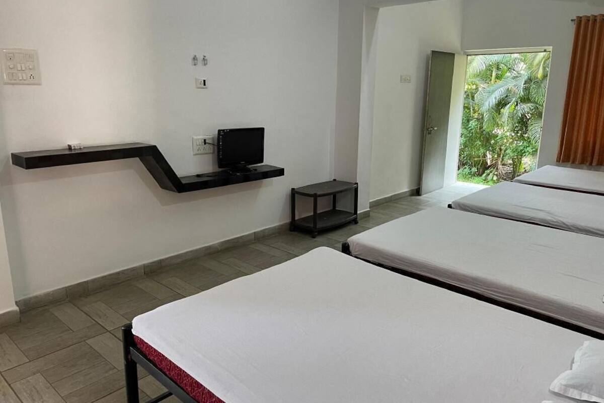 Quarto de hotel com duas camas, uma televisão em uma prateleira, uma pequena mesa e uma porta que dá para uma varanda com vegetação.