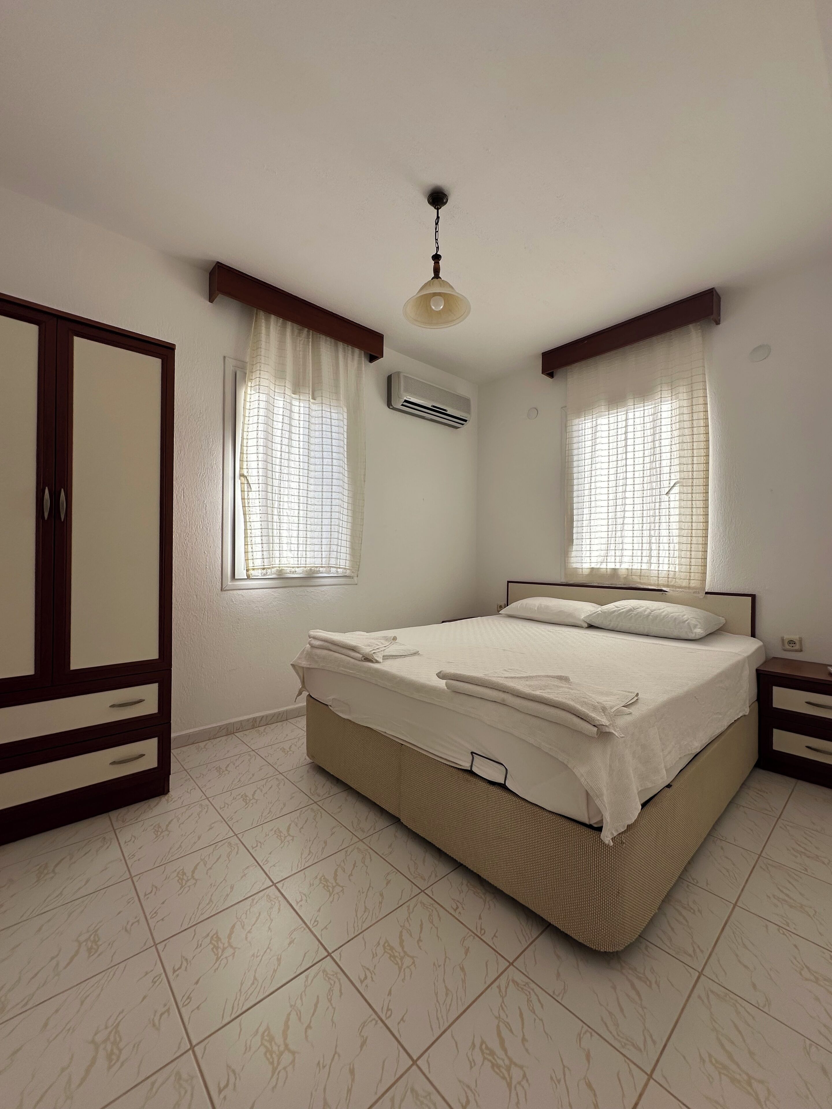 Quarto com cama, guarda-roupa, aparelho de ar condicionado e duas janelas com cortinas.