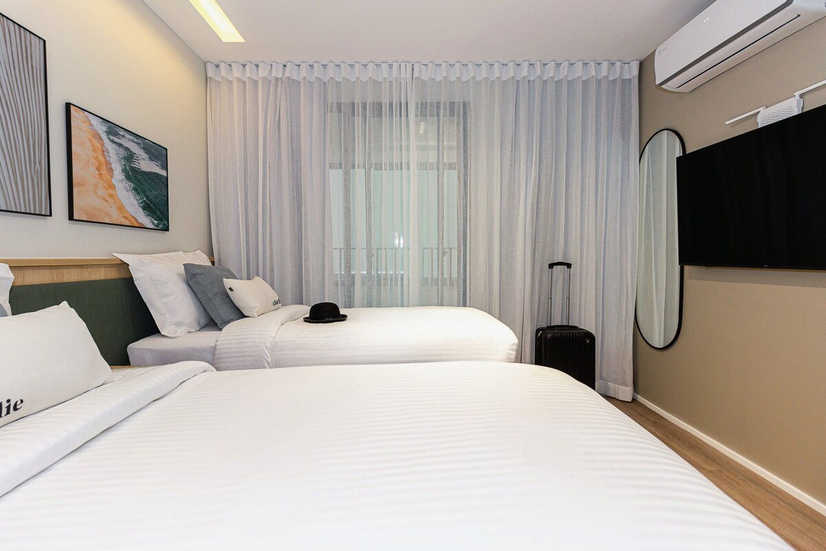 Quarto de hotel com duas camas, uma janela ampla com cortinas leves, ar condicionado, uma mala e uma TV de tela plana.