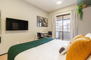 Quarto de hotel com cama, escrivaninha com cadeiras, mesinha com uma taça de vinho e vista da cidade por uma janela grande.
