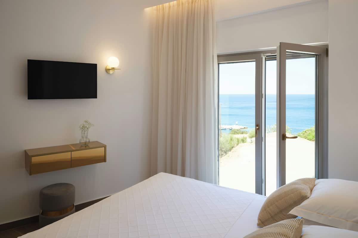 Quarto de hotel com cama, televisão montada, mesa de cabeceira com um vaso e vista para o mar através de uma porta de correr.