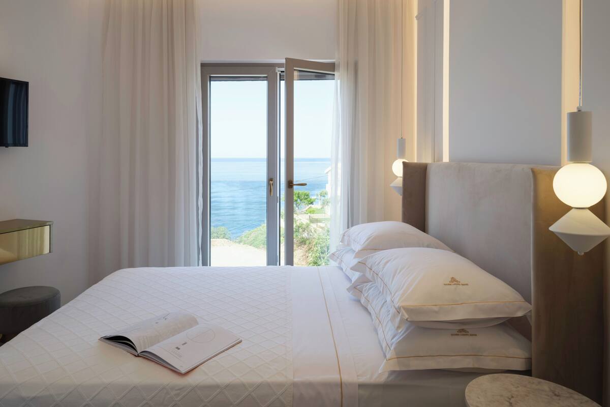 Quarto de hotel com cama, mesa de cabeceira, abajur e vista para o mar através de uma porta de vidro.