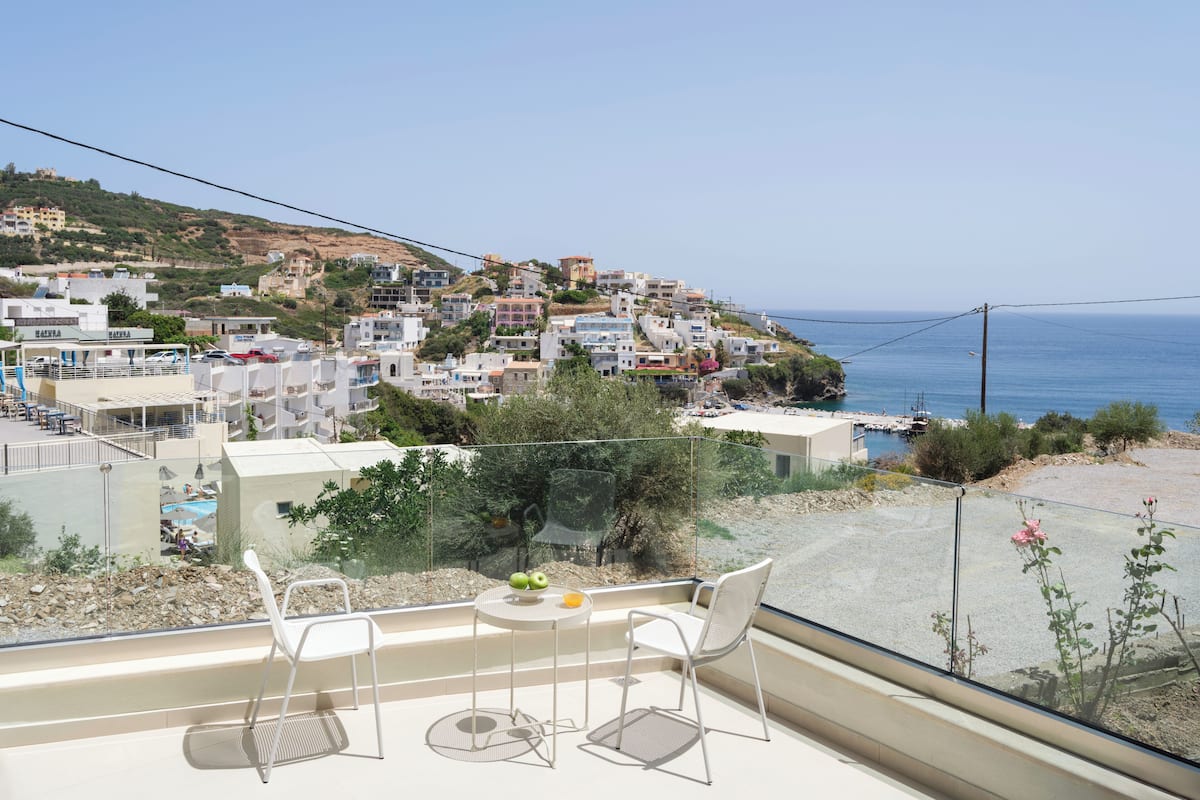 Varanda com mesa e cadeiras, vista para uma cidade costeira com edifícios e o mar.