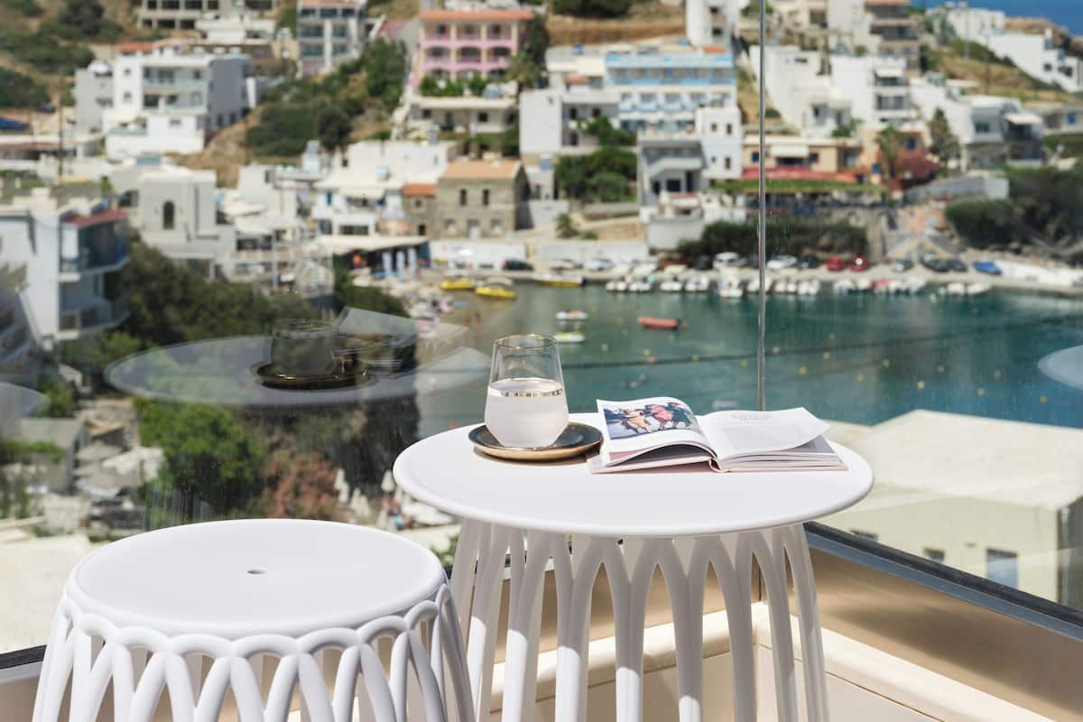 Varanda com mesa, um copo de água e uma revista. Na paisagem, uma vila costeira com edifícios e barcos.