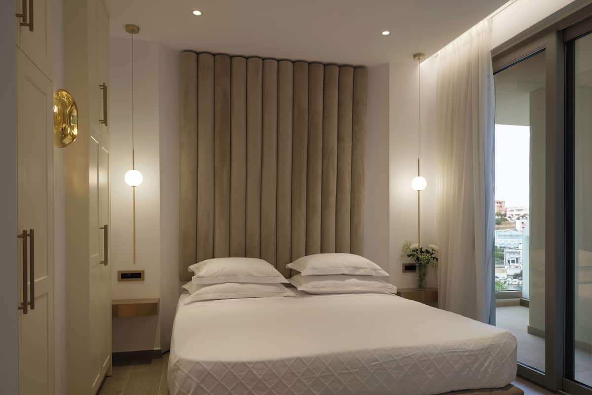 Quarto moderno com uma cama grande, painel de cabeceira vertical, abajures e vista da cidade através de uma porta deslizante.