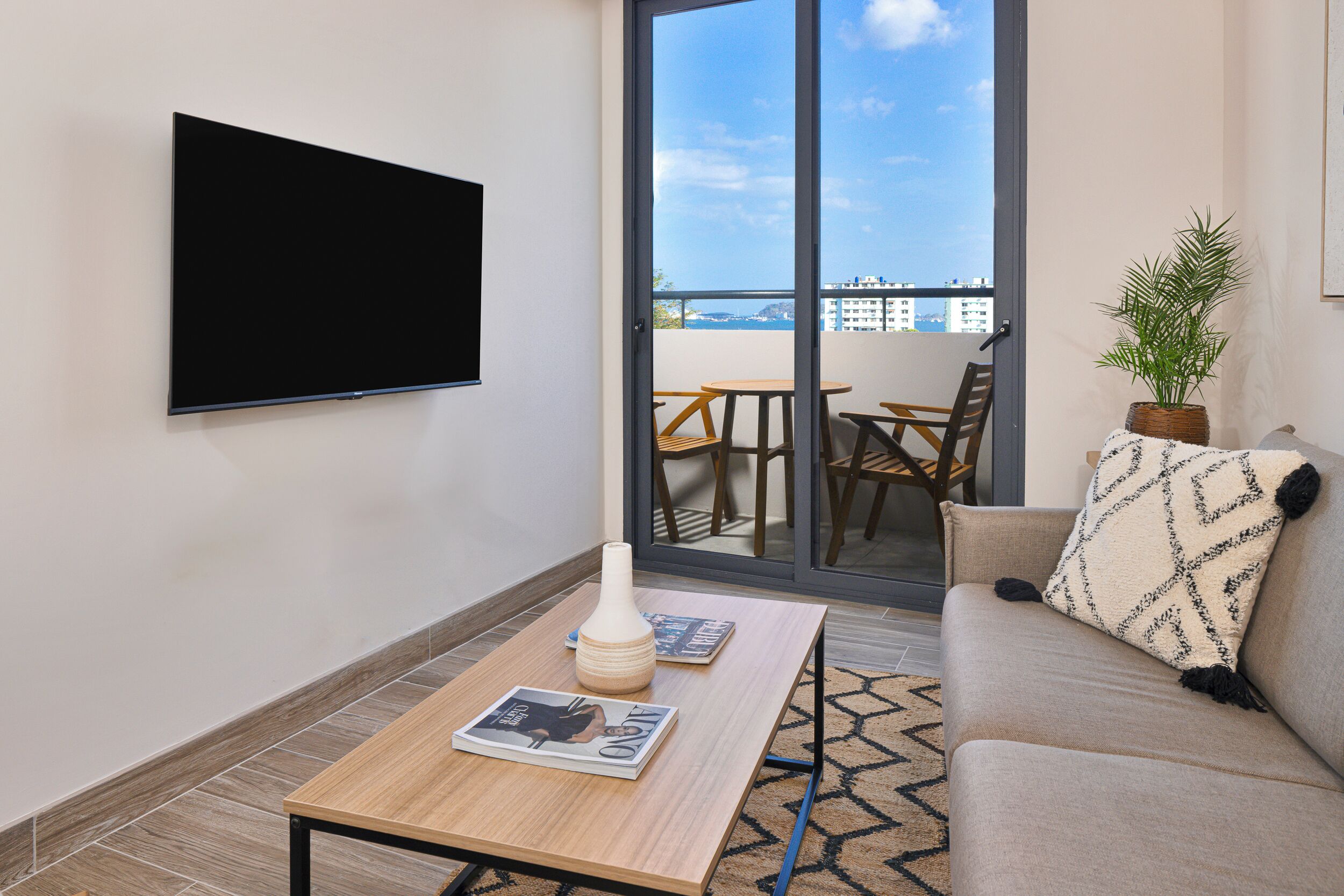 Uma sala de estar moderna com uma televisão de tela plana na parede, uma mesa de centro de madeira com revistas, um sofá bege com almofadas estampadas e um vaso de planta.