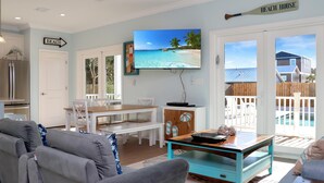 Sala de estar com TV exibindo uma cena de praia, placa com a inscrição "BEACH HOUSE" e vista para um deck de madeira e piscina.