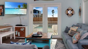 Sala de estar com televisão exibindo uma cena de praia, mesa de centro de madeira, sofá com almofadas estampadas e vista para uma casa através de portas de vidro.
