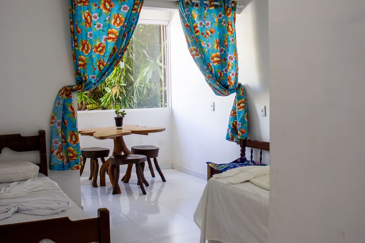 Quarto com cama, mesa, cadeiras e janela com cortinas.