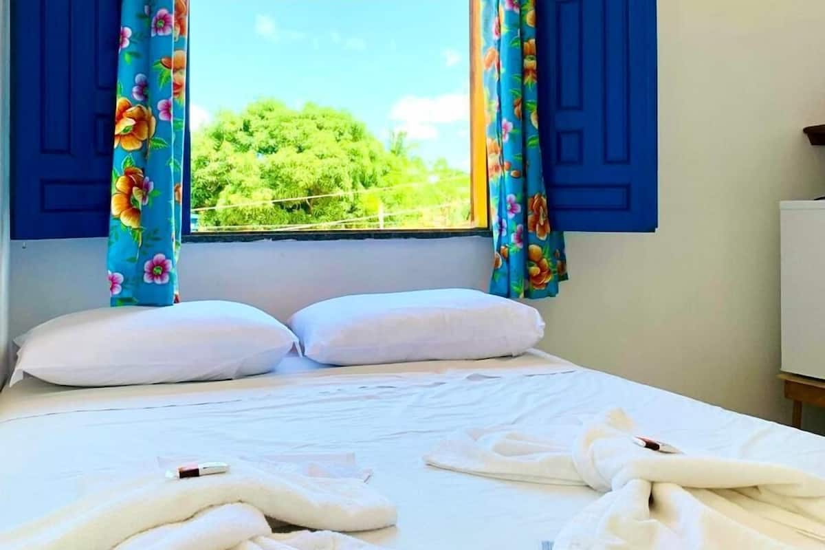 Quarto com cama, persianas azuis, cortinas florais e vista para o verde lá fora.