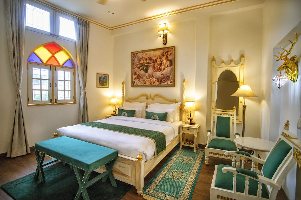 Quarto bem decorado com uma cama grande, mesas de cabeceira com luminárias, uma poltrona estofada verde e um espelho decorativo.
