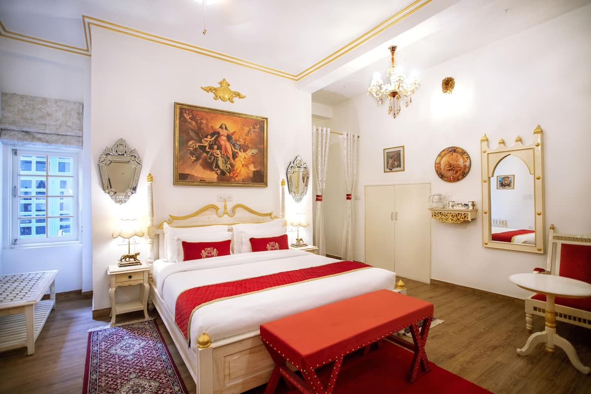 Quarto bem iluminado com uma cama grande, um banco vermelho, uma mesinha e um espelho.