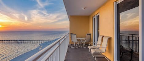 Uma varanda com vista para o oceano e um pôr do sol. Há duas cadeiras e uma mesa na varanda.