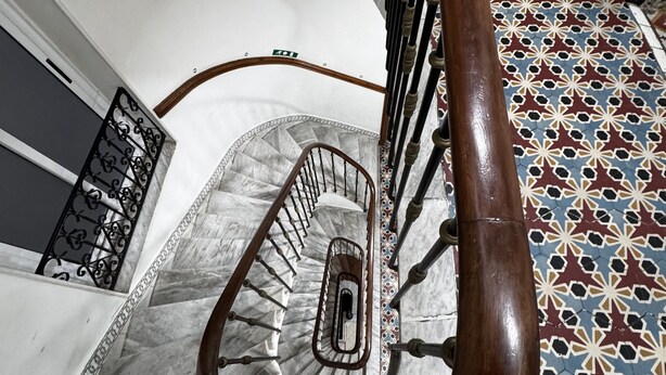 Uma escadaria de mármore com guarda-corpo de madeira e balaústres pretos, que leva a um patamar com piso estampado.