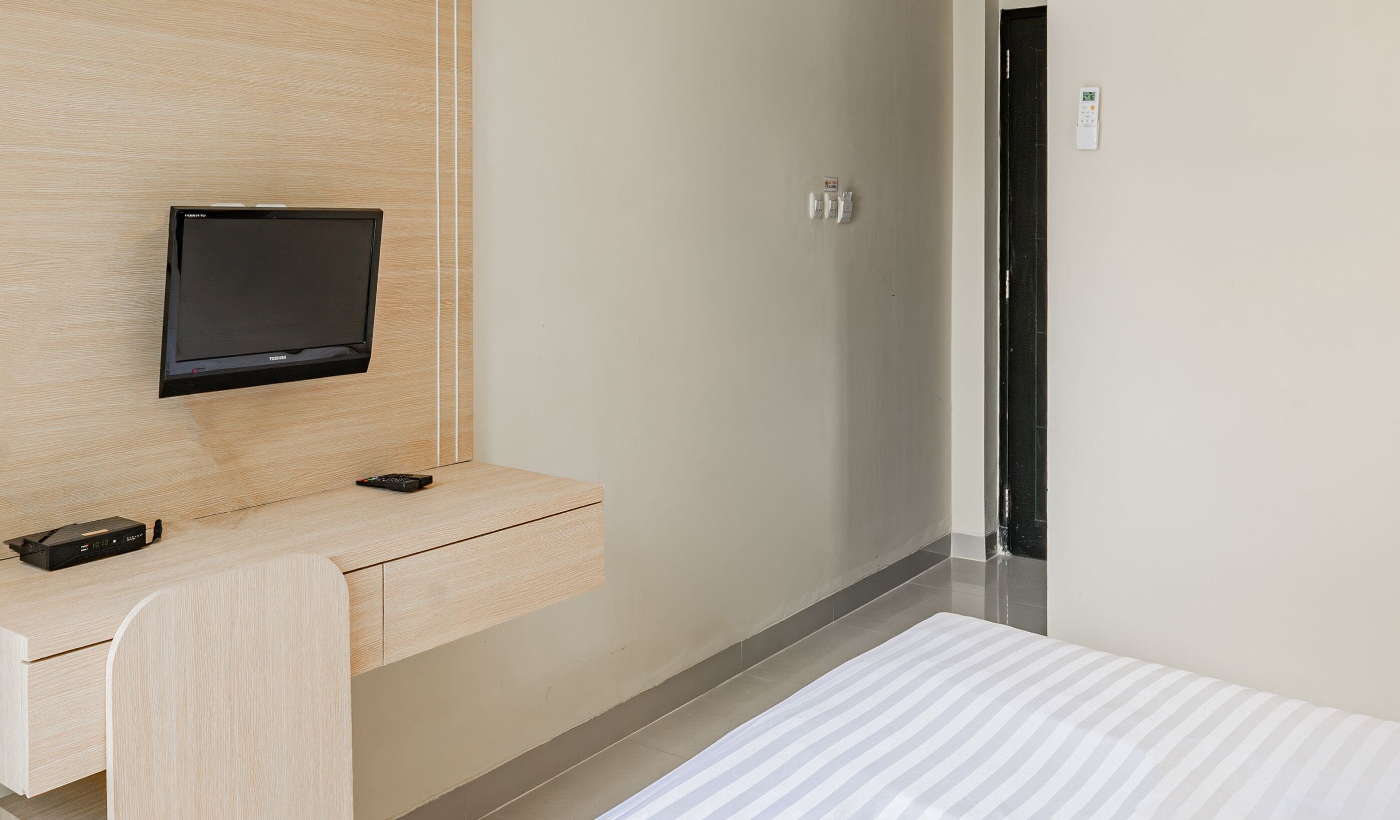 Quarto de hotel com televisão presa a uma parede revestida de madeira, uma pequena mesa com um controle remoto, uma cama com colcha listrada e uma porta que dá para outro quarto.