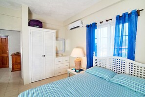 Quarto com cama, mesa de cabeceira com abajur, guarda-roupa e janela com cortinas azuis.