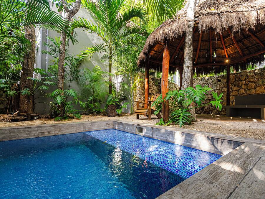 Piscina com borda azulejada, rodeada por plantas tropicais e uma estrutura de telhado de palha.