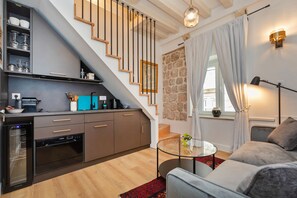 Uma cozinha moderna com forno embutido e máquina de lavar louça, uma escada com degraus de madeira, uma área de refeições com mesa de vidro e um espaço de estar acolhedor com sofá e luminária.
