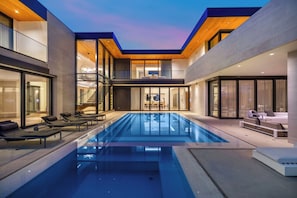 Piscina interior moderna com paredes de vidro, cadeiras de descanso e escadaria que leva aos níveis superiores.