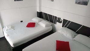 Quarto de hotel com duas camas, cada uma com roupa de cama branca e almofadas vermelhas. Possui uma cabeceira preta e branca, e uma pequena estante com um controle remoto e revistas.