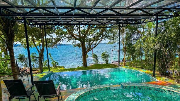 Uma piscina infinita circular com teto de vidro, rodeada por espreguiçadeiras e com vista para um corpo d'água.