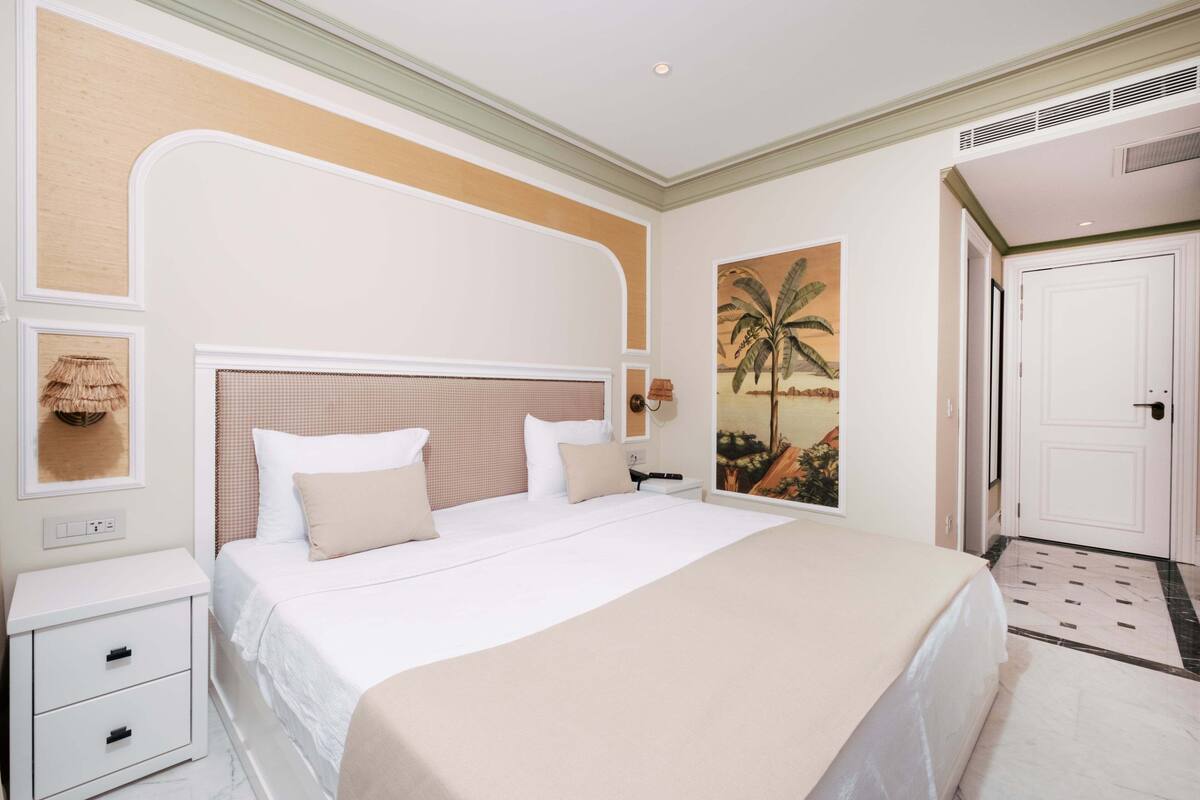 Quarto bem arrumado com uma cama grande, uma mesa de cabeceira, um abajur fixado na parede e um quadro de uma palmeira.