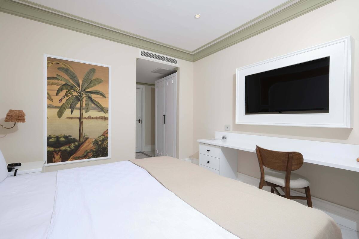 Um quarto com uma cama, uma escrivaninha, uma cadeira, uma televisão e um quadro de uma palmeira.