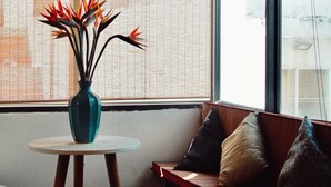 Um quarto com janela, um vaso com flores, uma mesa redonda e um banco com almofadas.