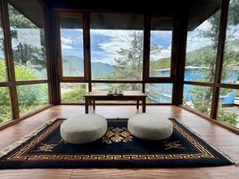 Um quarto com janelas amplas, uma mesa de madeira com xícaras e pires, dois travesseiros brancos e um tapete estampado.