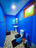 Casa de banho com paredes azuis, uma sanita branca, um lavatório e uma janela com vista para vegetação.