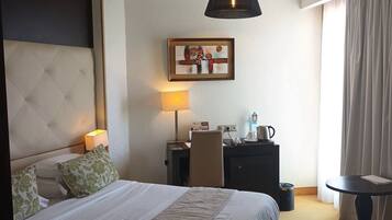 Quarto de hotel com uma cama grande, uma escrivaninha com luminária, uma cadeira, uma mesa pequena e um quadro na parede.