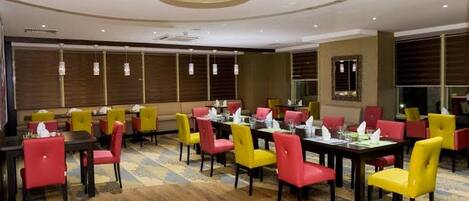 Uma sala de jantar de hotel com várias mesas e cadeiras, um tapete estampado e um lustre grande.