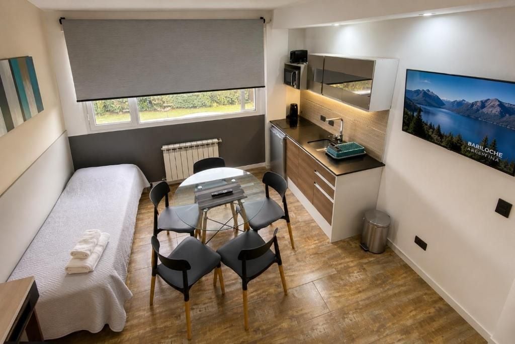 Quarto compacto com cama, mesa de jantar e cadeiras, pequeno espaço de cozinha e televisão na parede.