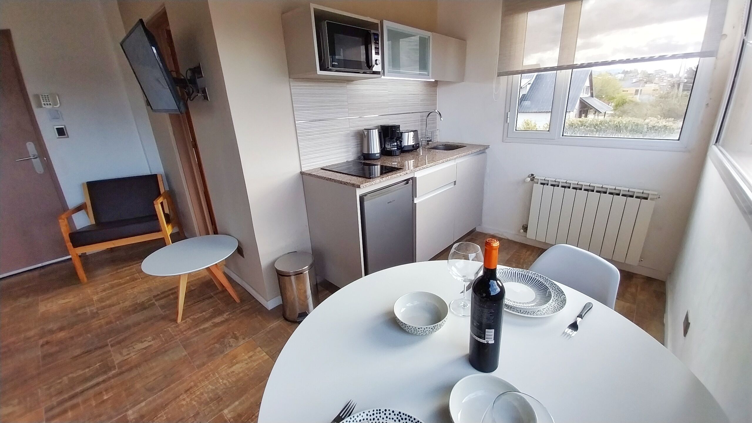 Cozinha compacta equipada com micro-ondas, máquina de café e pequena geladeira. Há uma mesa de jantar com pratos, uma garrafa de vinho e uma cadeira. O quarto possui uma janela com vista para o exterior.