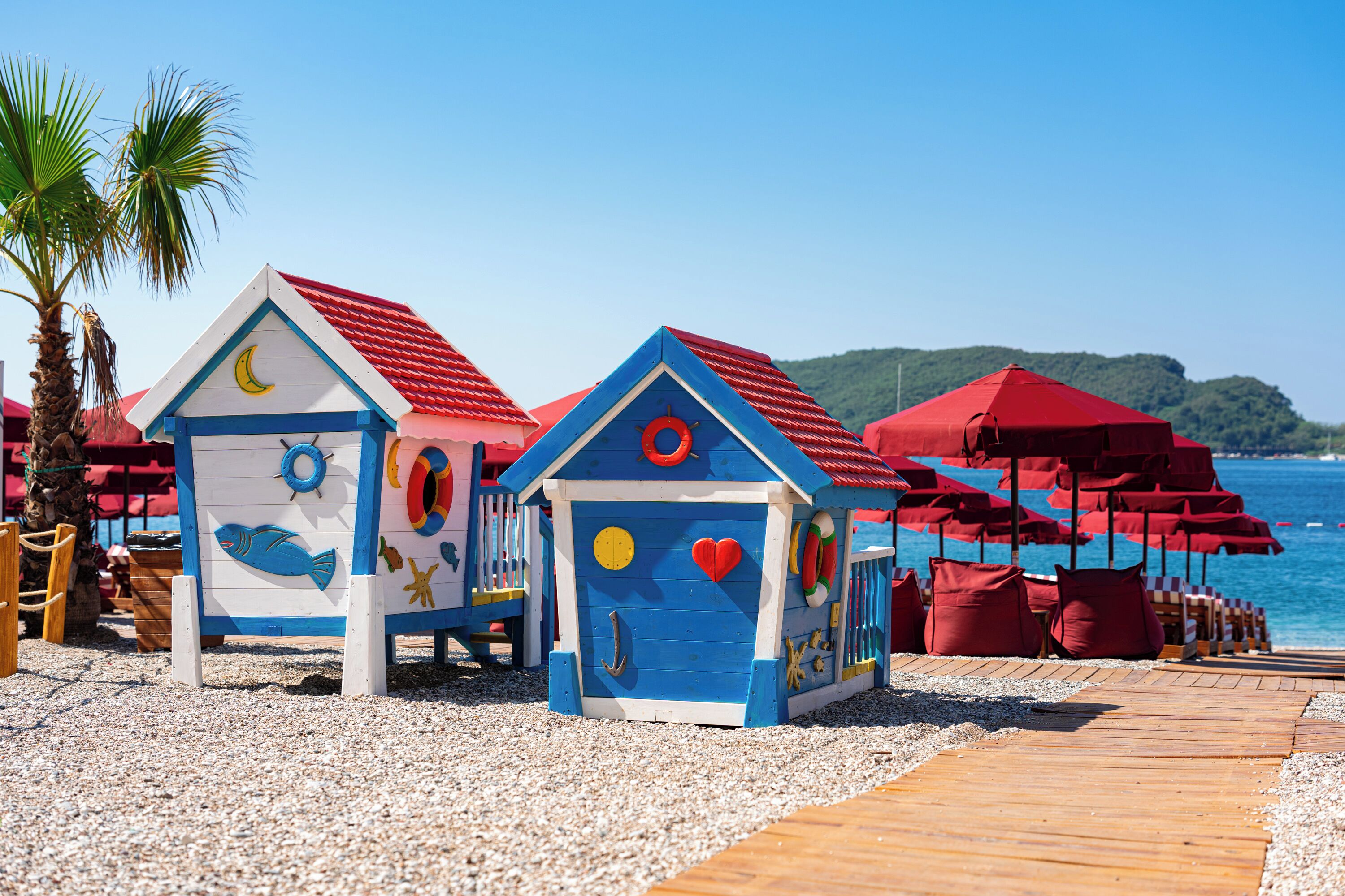 Bungalôs de praia com telhado vermelho e base azul, um deck de madeira e guarda-sóis vermelhos.