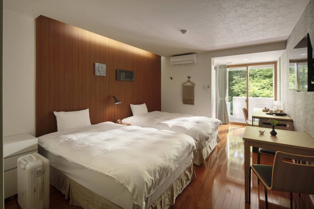 Quarto de hotel com duas camas, cabeceira de madeira, uma mesinha com uma planta e vista para o verde pela janela.