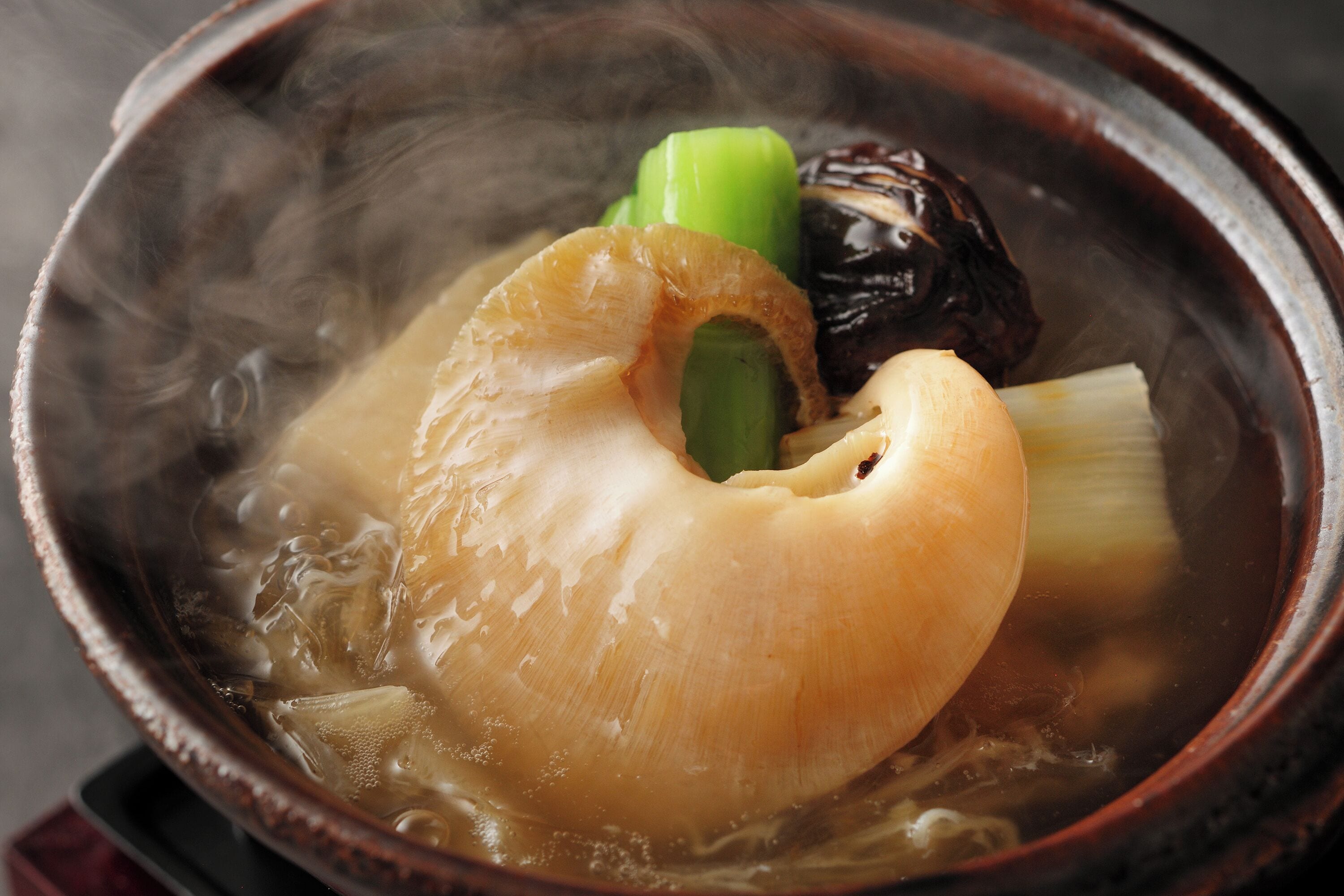 Uma tigela de sopa com uma amêijoa grande, um legume verde e um item escuro, possivelmente um cogumelo, em uma tigela de cerâmica.